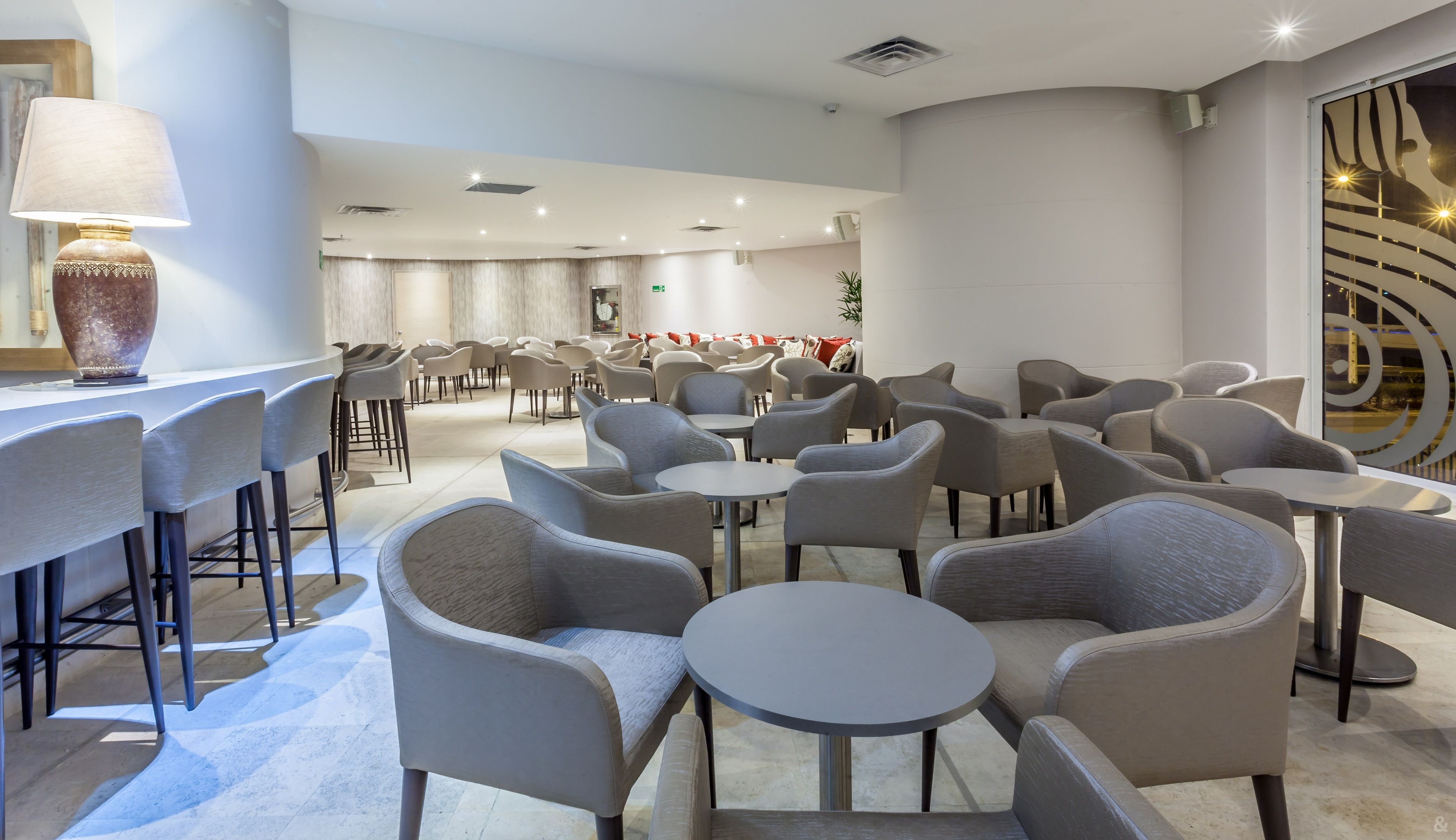 Um amplo hall de hotel com várias áreas de estar, incluindo mesas redondas e cadeiras estofadas, e um grande vaso decorativo sobre uma mesa.