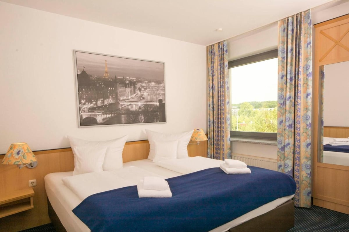 Quarto de hotel com cama, mesa de cabeceira, luminária, janela com cortinas e uma fotografia da paisagem urbana na parede.