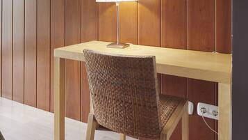 Uma mesa de madeira com cadeira de fibra natural, uma televisão na parede e um abajur.