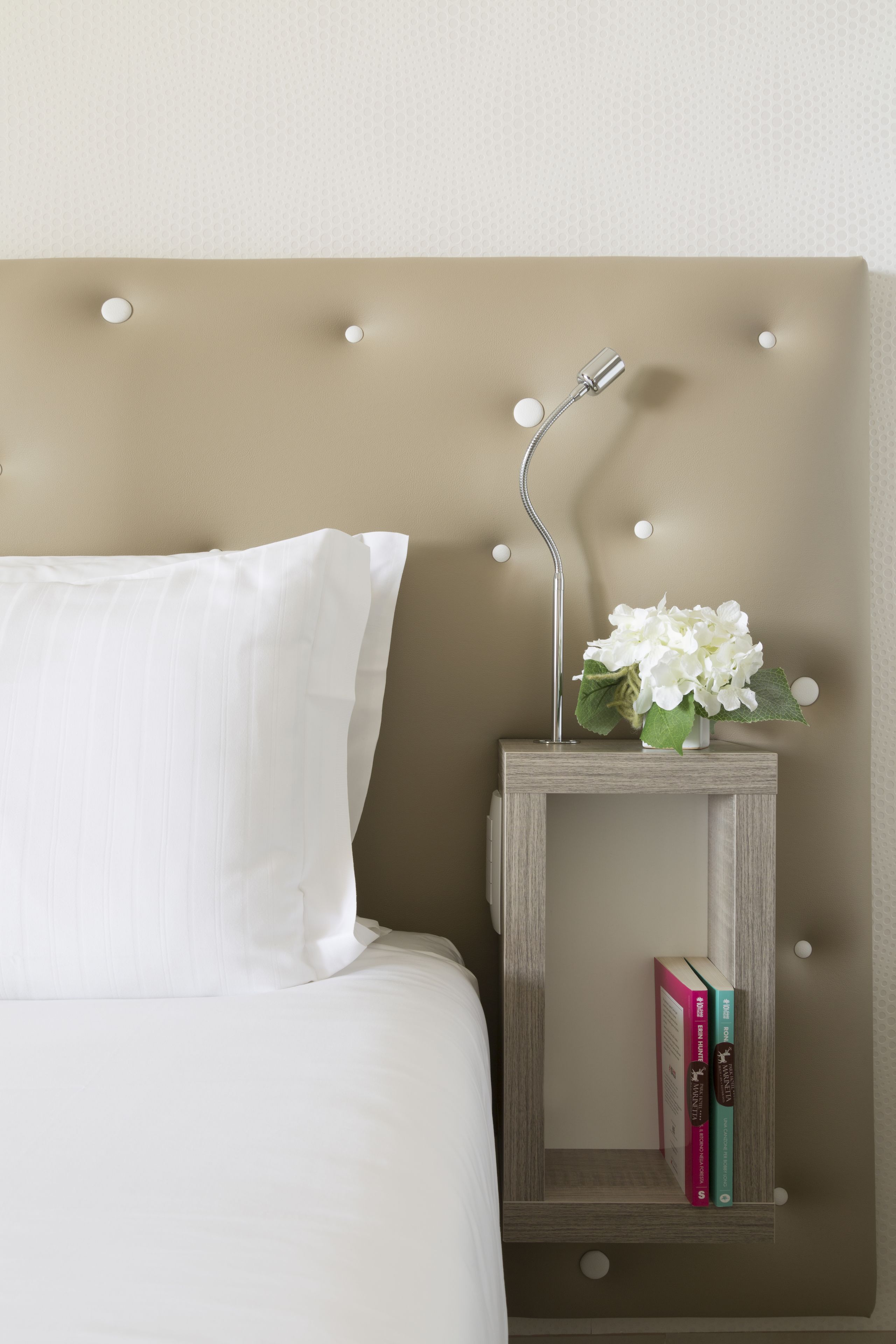 Cama bem arrumada com travesseiro branco e mesa de cabeceira de madeira com abajur de metal, um vaso com flores brancas e dois livros.
