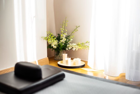 Quarto com piso de madeira, um objeto preto sobre uma mesa e um vaso com flores brancas e velas.