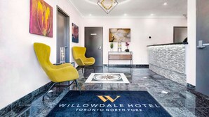 Um saguão de hotel com um balcão de recepção, duas cadeiras amarelas, uma grande pintura abstrata e piso de mármore.
