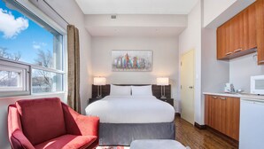 Quarto de hotel com uma cama, uma cadeira vermelha, uma mesa pequena, um quadro com vista para o horizonte da cidade, uma janela com cortinas e uma cozinha compacta.