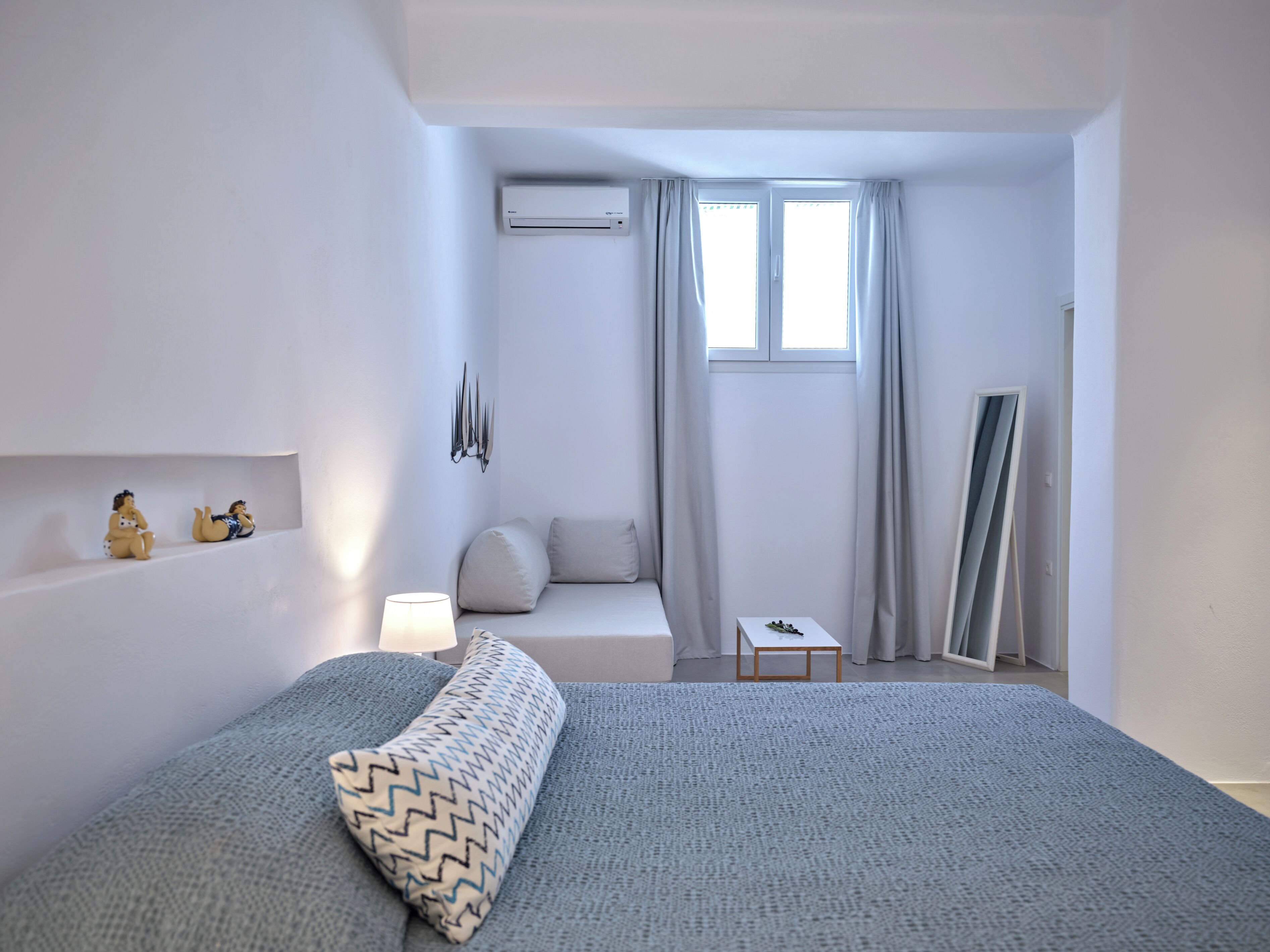 Quarto moderno com cama, mesa de cabeceira, pequena mesa, janela com cortinas, ar condicionado e obras de arte na parede.