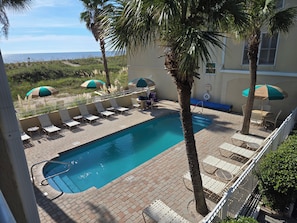 Área da piscina com espreguiçadeiras e guarda-sóis, vista desobstruída do oceano e um edifício com fachada bege.