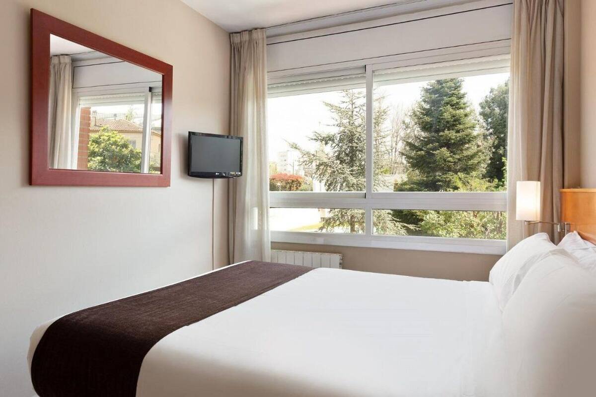 Quarto de hotel com cama, televisão e janela com vista para árvores e edifícios.