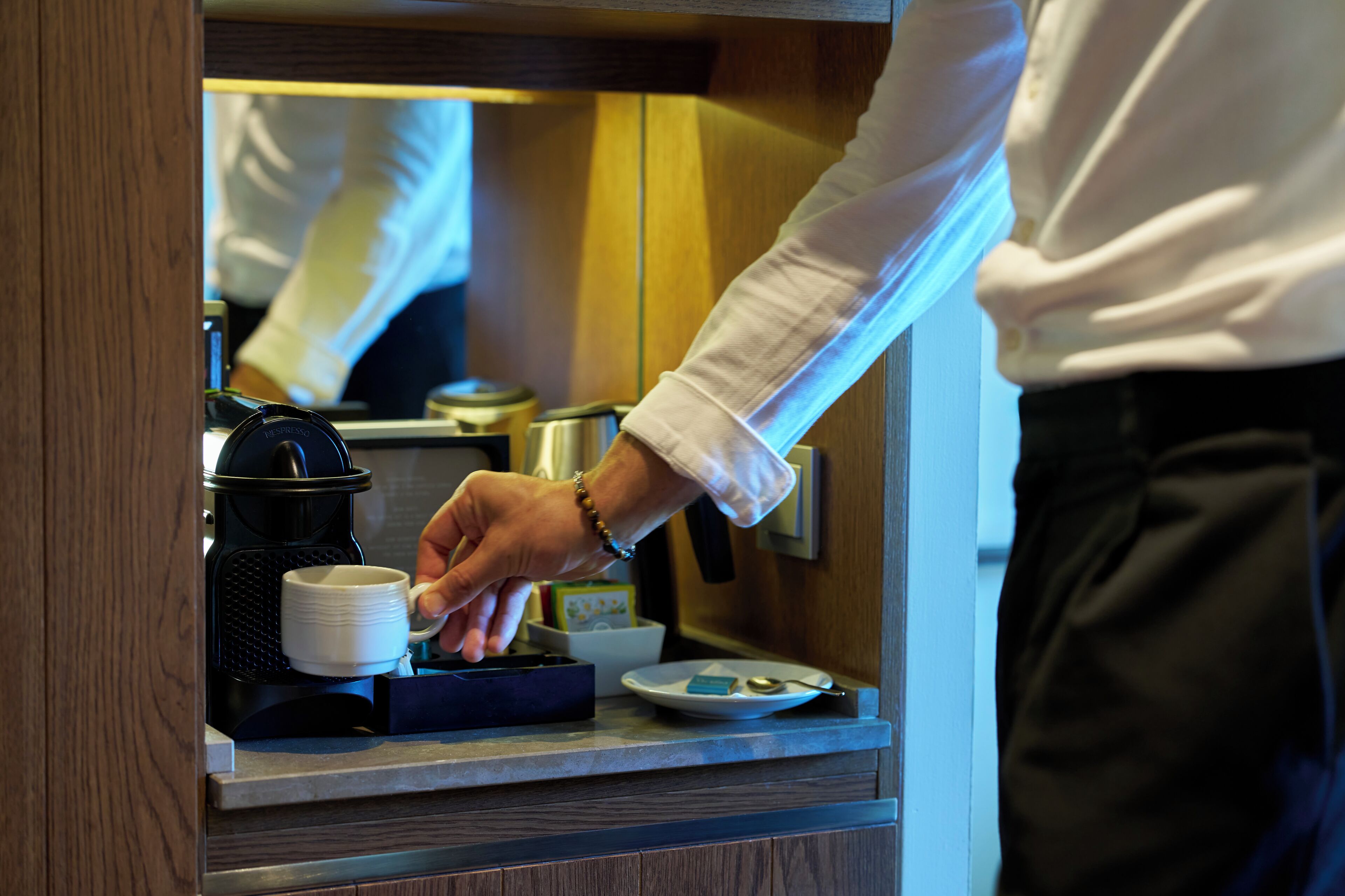 Uma pessoa manuseando uma máquina de café, com uma xícara e um prato com colher sobre o balcão.