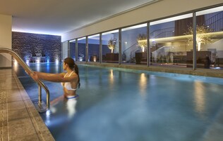 Piscina interior moderna com barreira de vidro, uma mulher de fato de banho e vista para um edifício com escadas e plantas.