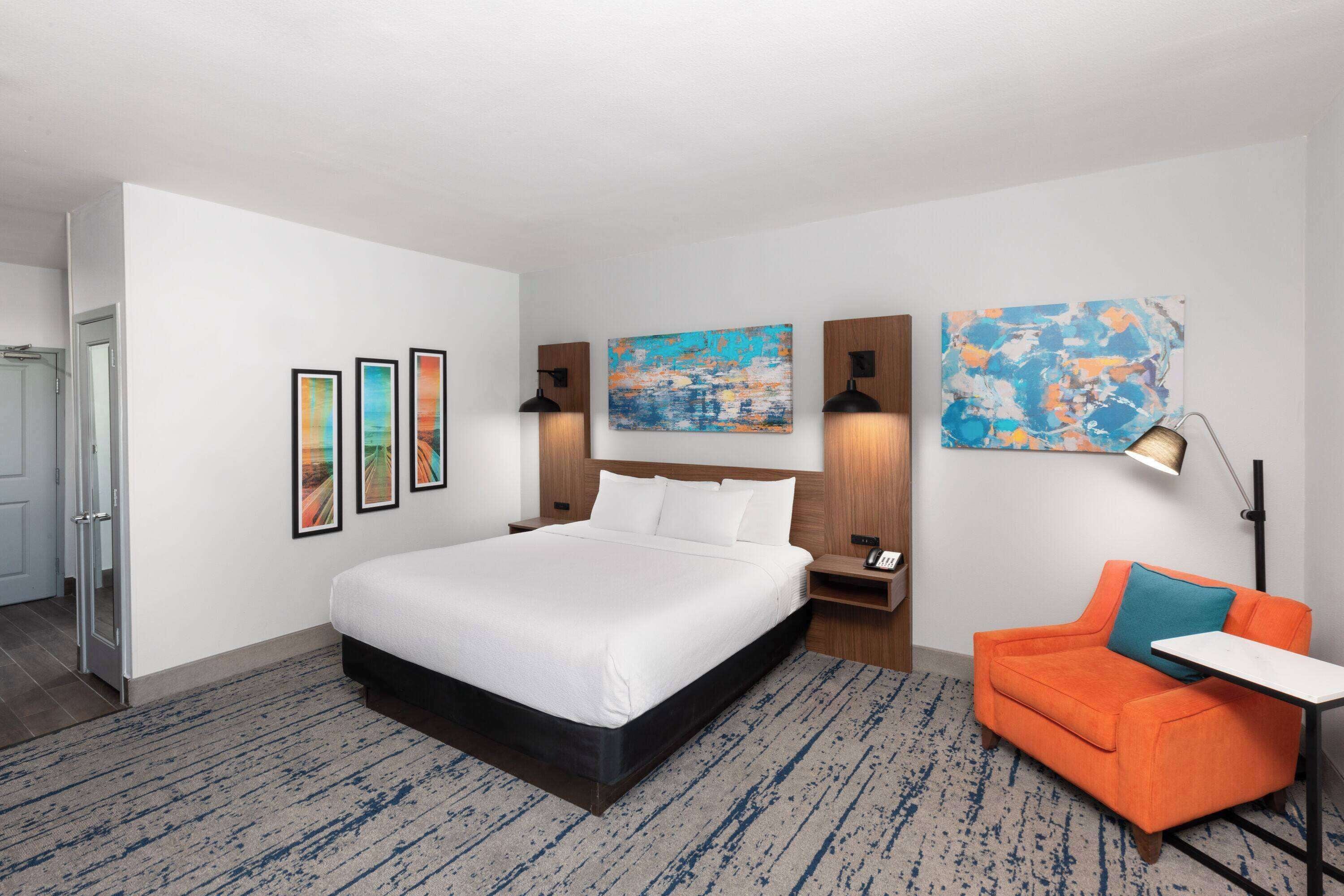 Quarto de hotel com uma cama grande, uma mesinha de cabeceira, uma escrivaninha, uma poltrona laranja e duas pinturas abstratas nas paredes.