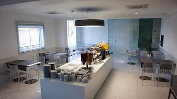 Quarto de hotel moderno com uma ilha central, área de refeições com cadeiras e mesas, e uma grande janela com persianas.