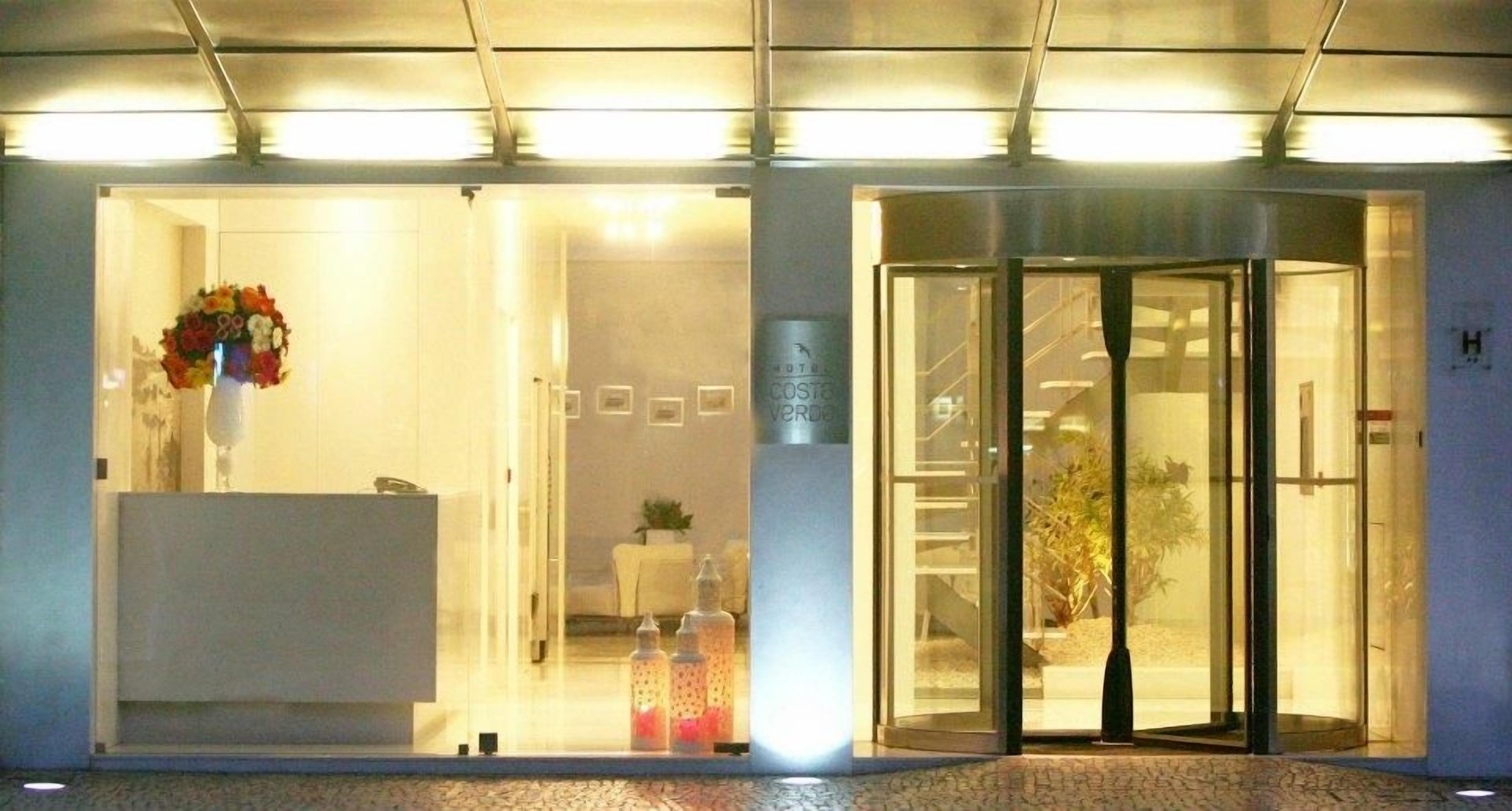 Entrada de hotel com balcão de recepção, porta de vidro e área interna com planta e espaço para sentar.