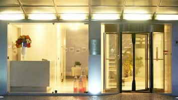 Entrada de hotel com balcão de recepção, porta de vidro e área interna com planta e espaço para sentar.