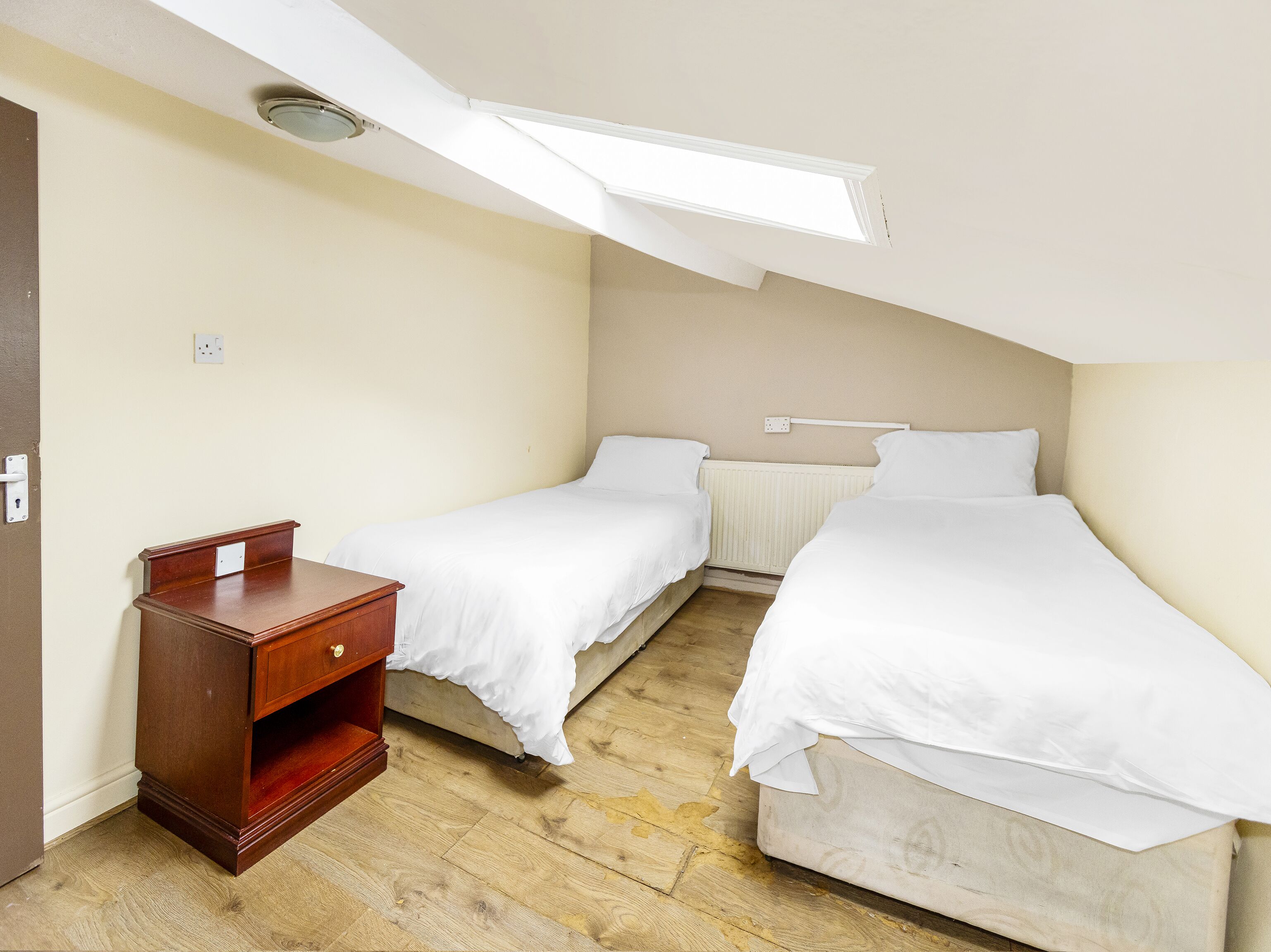 Quarto pequeno e limpo com duas camas de solteiro, uma mesa de cabeceira de madeira, um radiador e um clarabóia.
