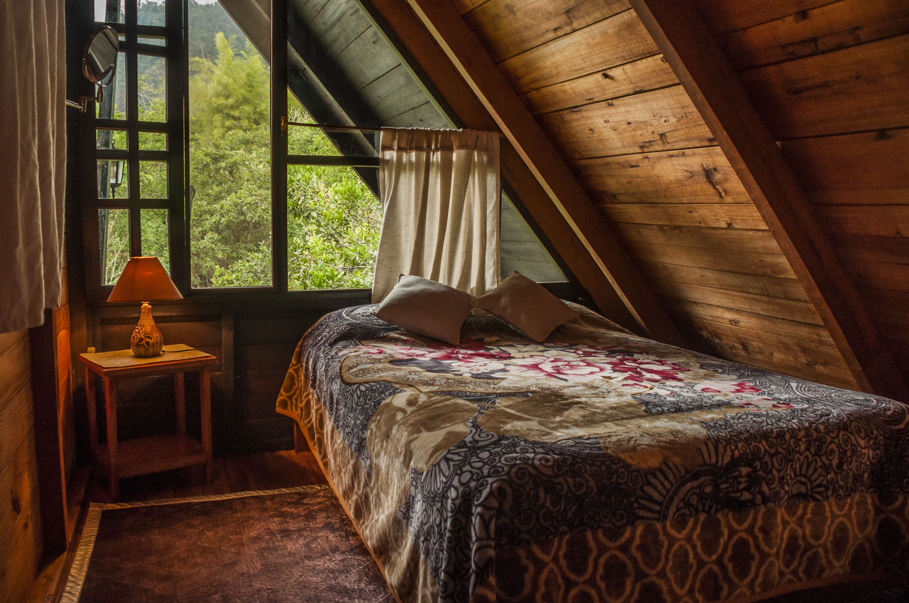 Um quarto de sótão acolhedor, com teto inclinado de madeira, uma janela grande com cortinas, uma cama com roupa de cama estampada e uma pequena mesa com abajur.
