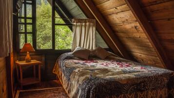 Um quarto de sótão acolhedor, com teto inclinado de madeira, uma janela grande com cortinas, uma cama com roupa de cama estampada e uma pequena mesa com abajur.