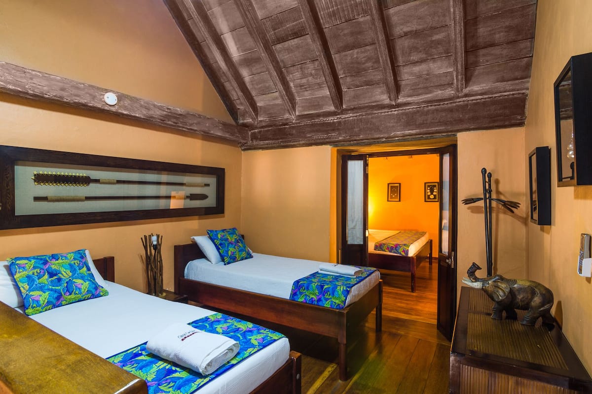 Quarto de hotel com duas camas, vigas de madeira no teto, um quadro emoldurado acima das camas e uma pequena estátua sobre uma mesa.