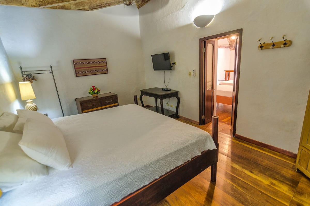 Quarto com cama, escrivaninha, televisão e porta para outro espaço.