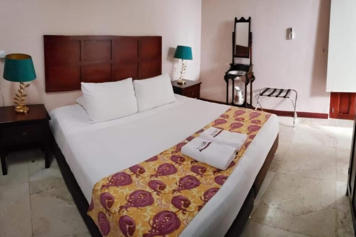Quarto de hotel com cama, duas mesas de cabeceira, um criado-mudo, um espelho e um banquinho.