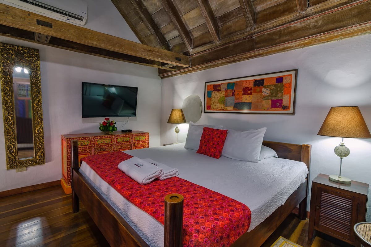 Quarto de hotel com teto de madeira, uma cama grande com colcha vermelha e branca, uma televisão na parede, um espelho decorativo e um colcha colorida pendurada sobre a cama.