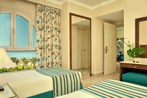 Quarto de hotel com duas camas, uma janela grande com vista para palmeiras, um banheiro com espelho e uma pequena escrivaninha com luminária.