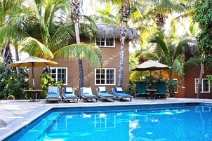 Uma área junto à piscina com espreguiçadeiras, guarda-sóis e um edifício com telhado de palha.