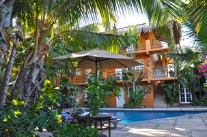 Área da piscina com espreguiçadeira e guarda-sol, cercada por vegetação exuberante e um edifício com varanda.
