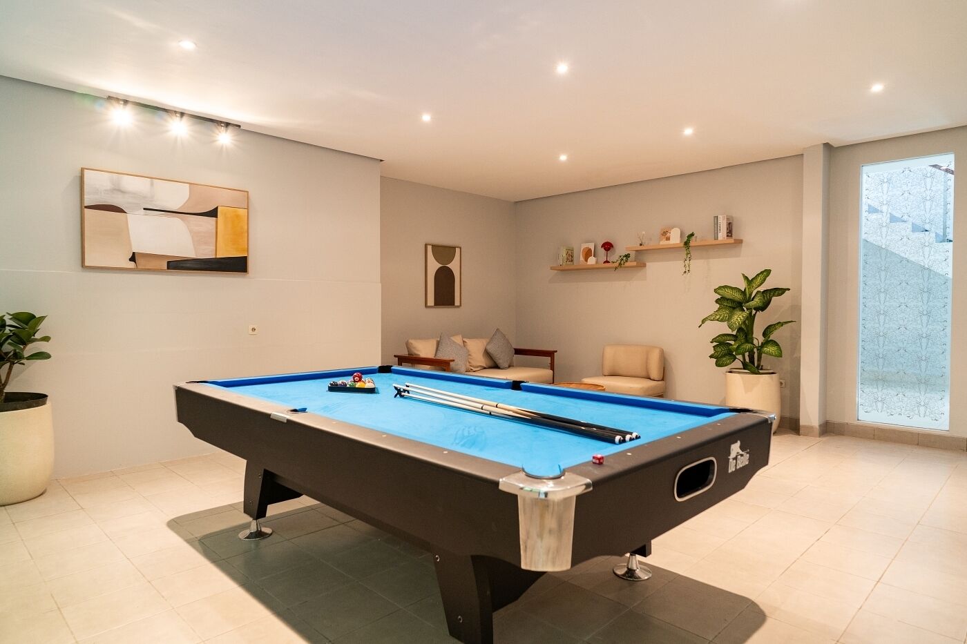 Uma mesa de bilhar em um ambiente moderno, com um sofá, uma planta em vaso e uma janela com vista.