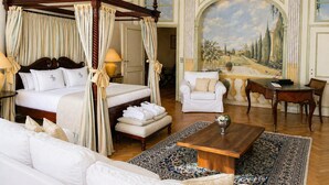 Uma cama com dossel e cortinas, um sofá branco, uma mesa de centro de madeira e uma grande pintura de paisagem na parede.