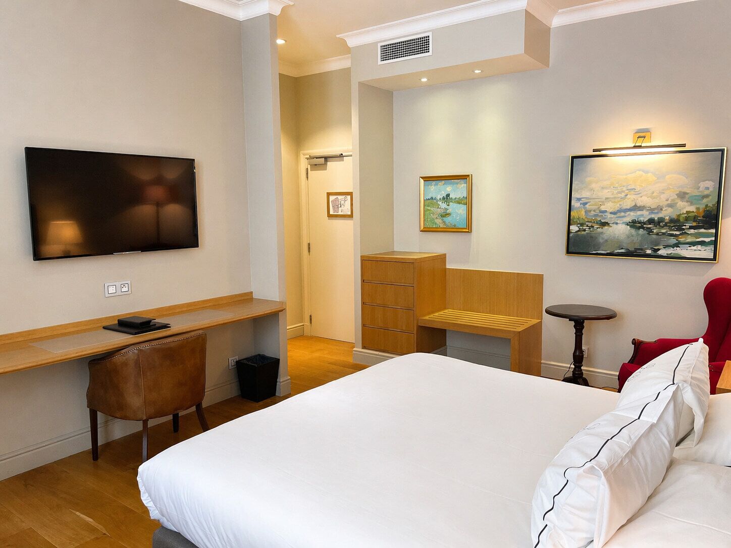 Quarto de hotel com cama, escrivaninha com cadeira, televisão, quadro e uma pequena mesa.