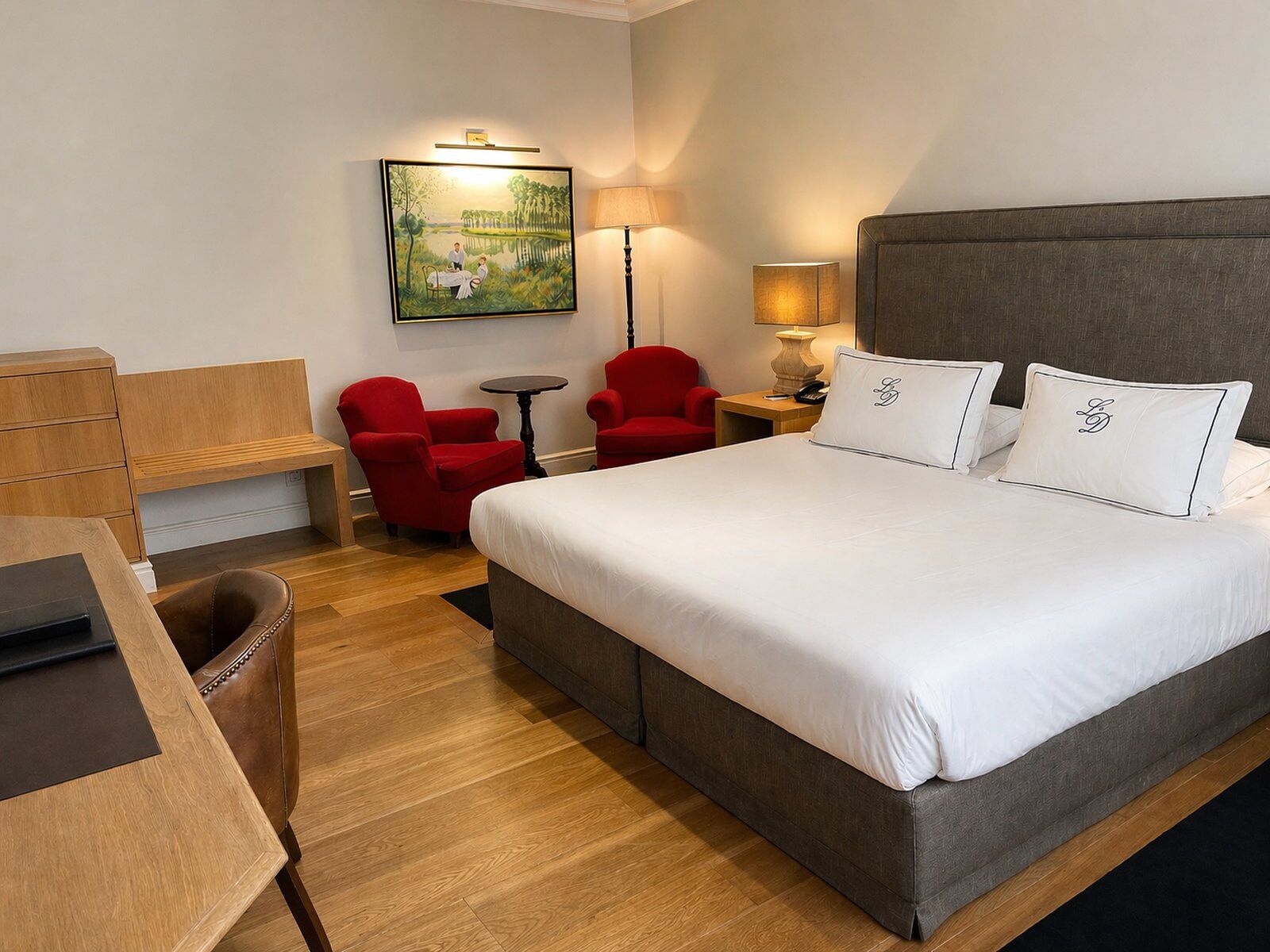 Quarto de hotel com uma cama grande, duas poltronas vermelhas, uma escrivaninha e um quadro na parede.