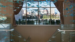 Um hall de hotel moderno com uma escadaria de vidro, vista da cidade através de amplas janelas e elementos decorativos pendentes.