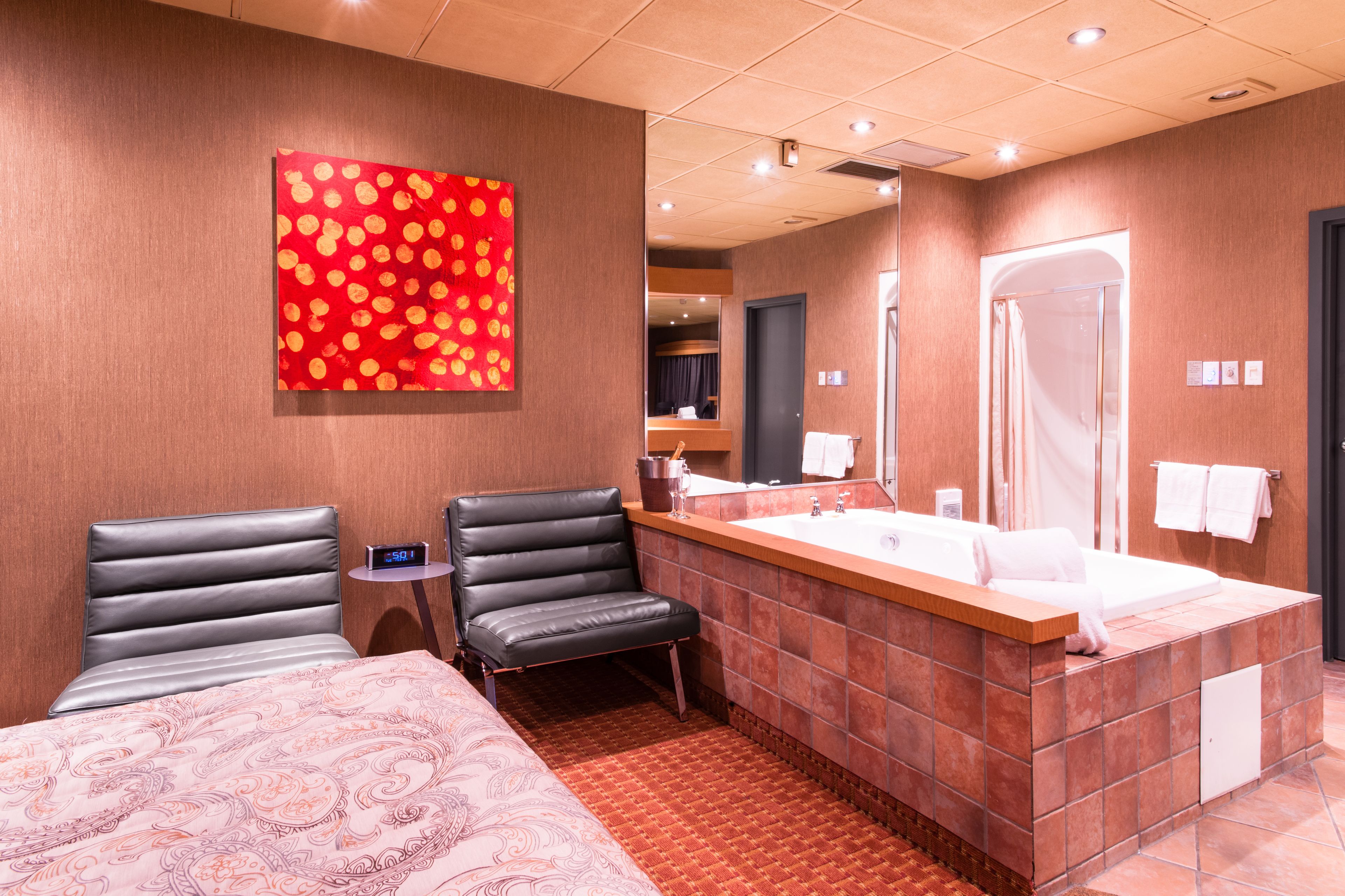 Quarto de hotel com cama, duas cadeiras, uma mesinha, um espelho grande, pia e área de chuveiro.