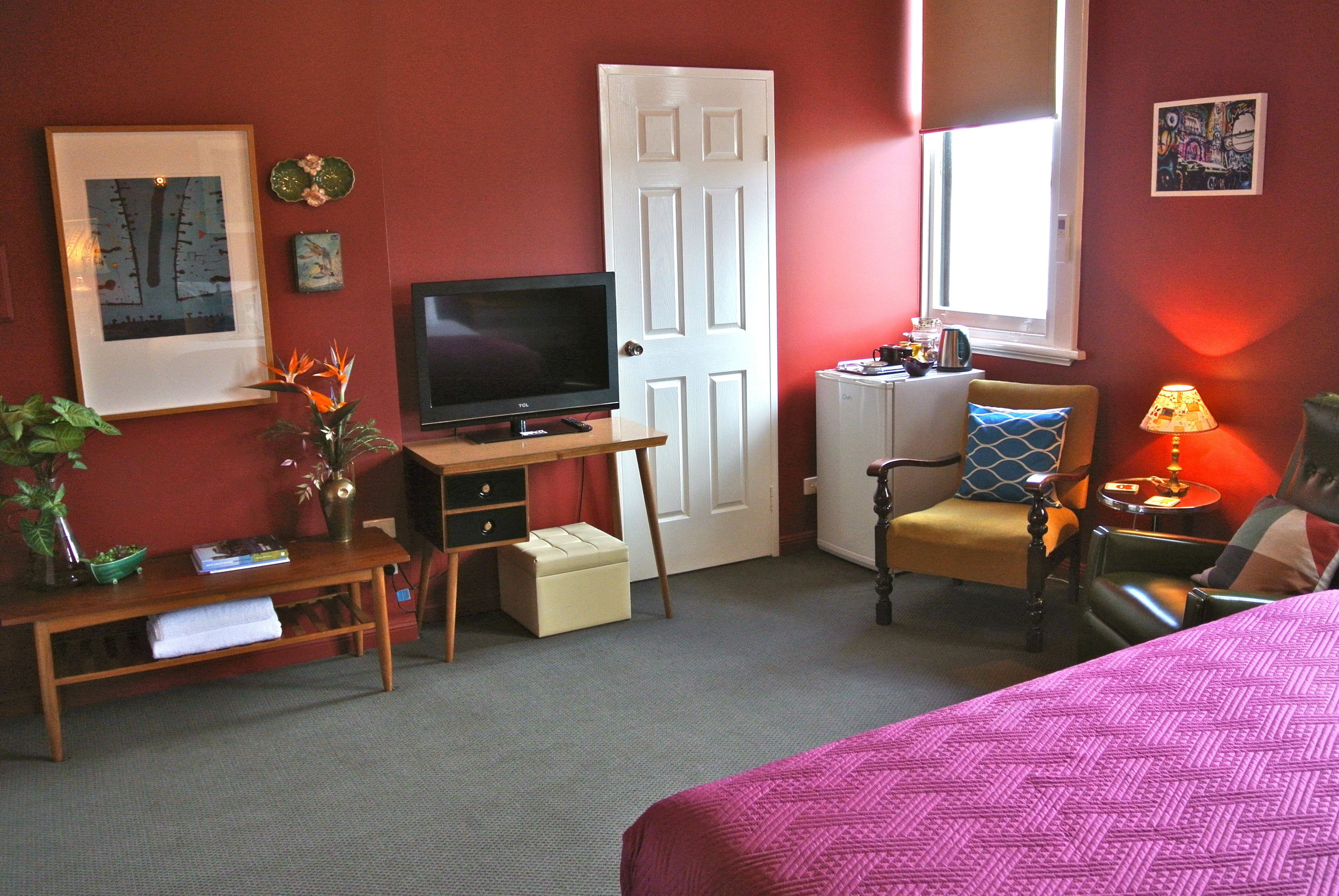 Quarto de hotel com cama, escrivaninha com televisão, cadeira, abajur e janela.