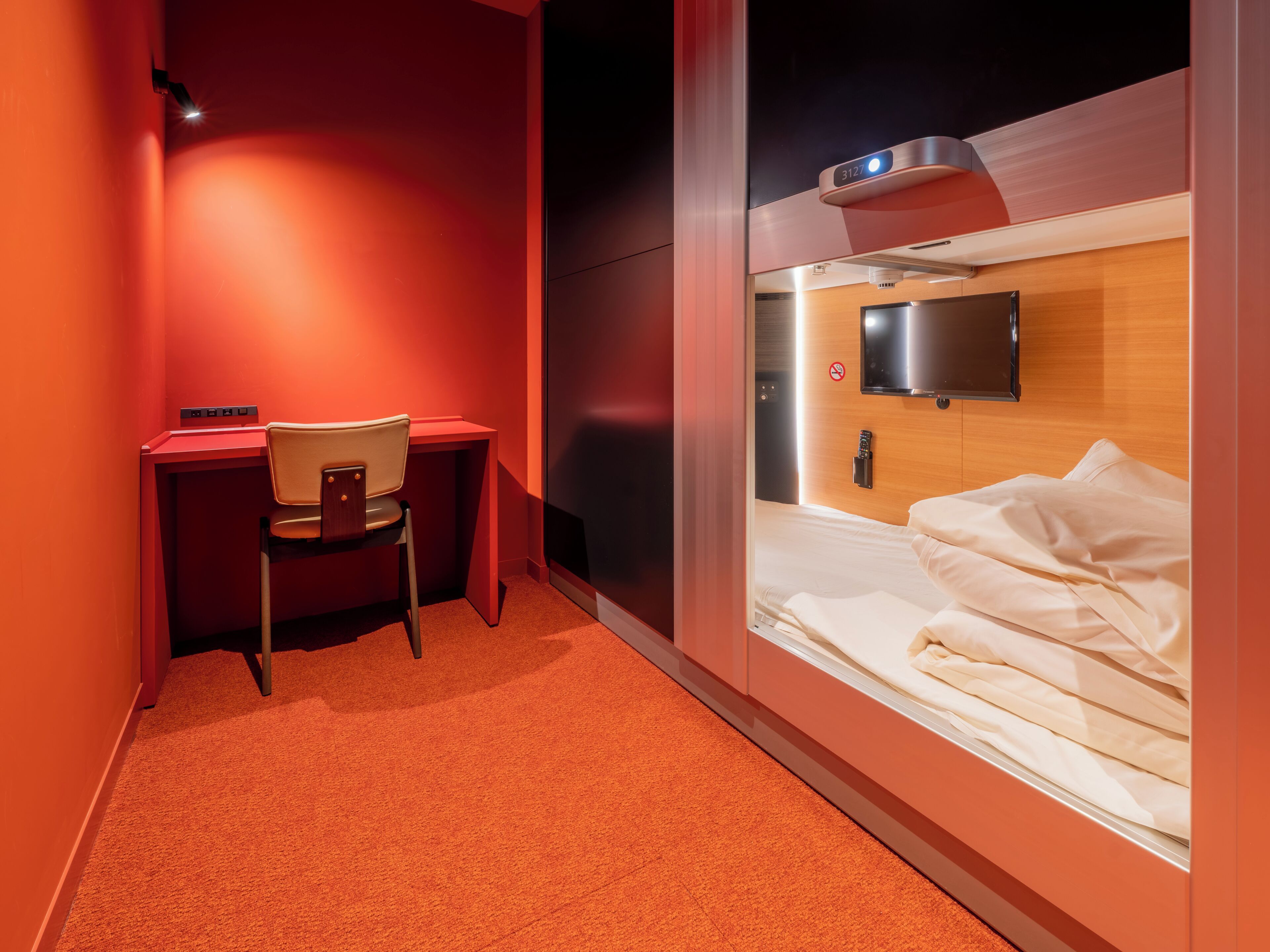 Quarto de hotel com cama, escrivaninha, cadeira, televisão e parede vermelha.