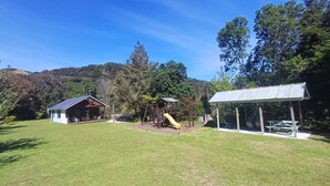 Uma área gramada com um parquinho, um pequeno edifício com telhado vermelho e um pavilhão coberto.