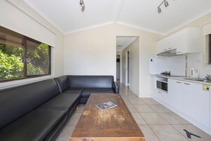 Cozinha moderna com armários brancos, forno embutido e máquina de café. Há um sofá de couro preto e uma mesa de centro de madeira com uma revista.