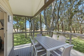 Espaço coberto ao ar livre com mesa e cadeiras, com vista para uma área gramada e árvores.