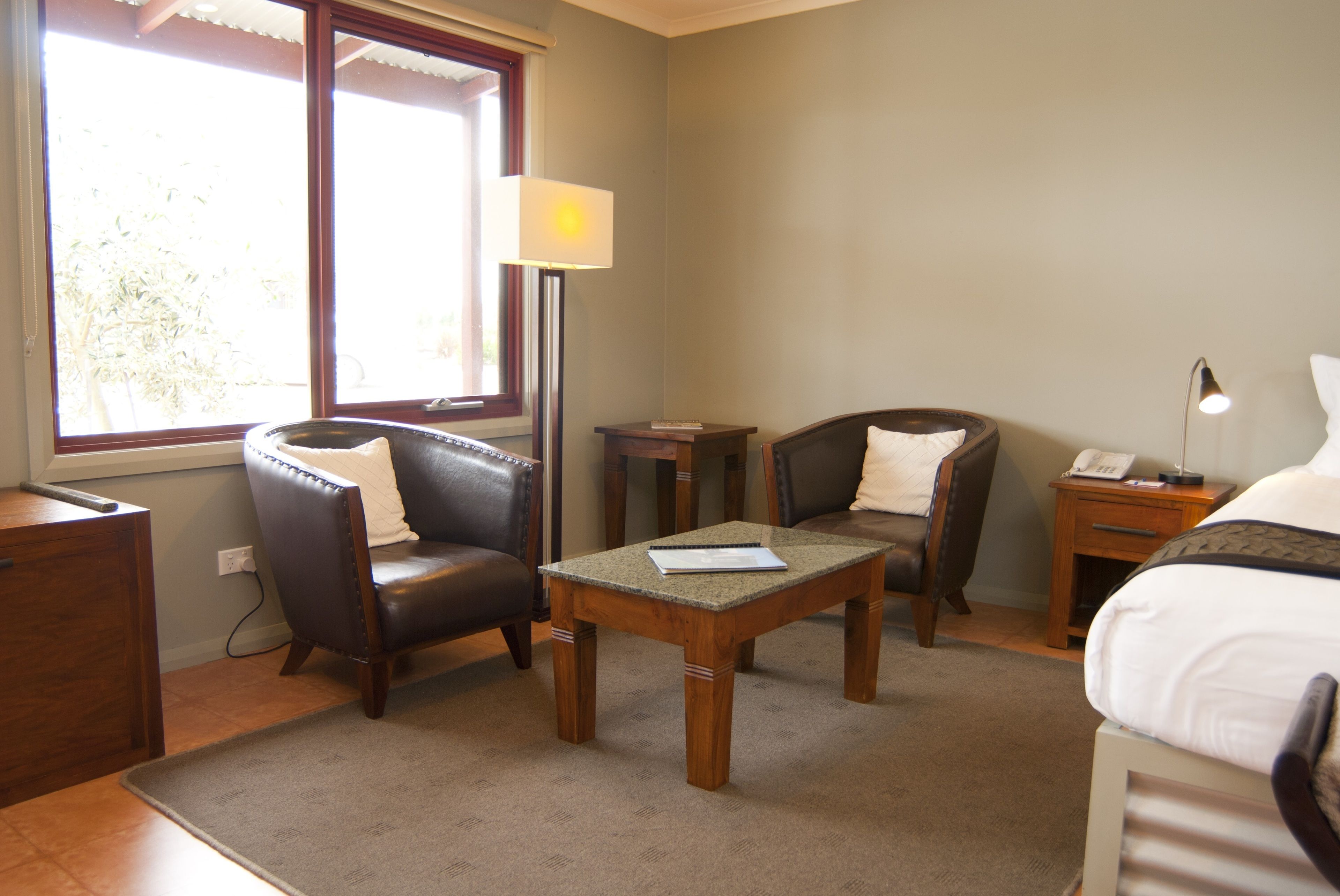 Quarto de hotel com cama, duas cadeiras, uma mesa pequena, abajur e janela com persianas.