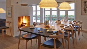 Uma sala de jantar com uma mesa comprida, cadeiras e luminárias pendentes. Uma lareira é visível ao fundo.