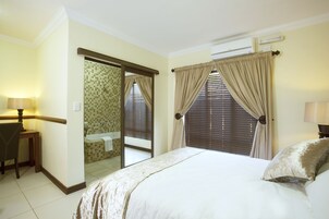 Quarto de hotel com cama, escrivaninha, espelho e banheiro visível através de uma porta aberta.