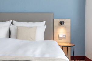 Cama bem arrumada com lençóis brancos e cabeceira de madeira. Um pequeno criado-mudo de madeira com uma luminária fixada na parede fica ao lado da cama.