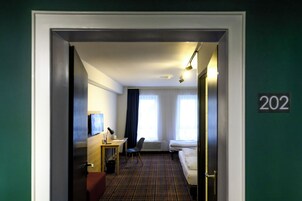 Quarto de hotel com cama, escrivaninha, cadeira, televisão e janela com cortinas.