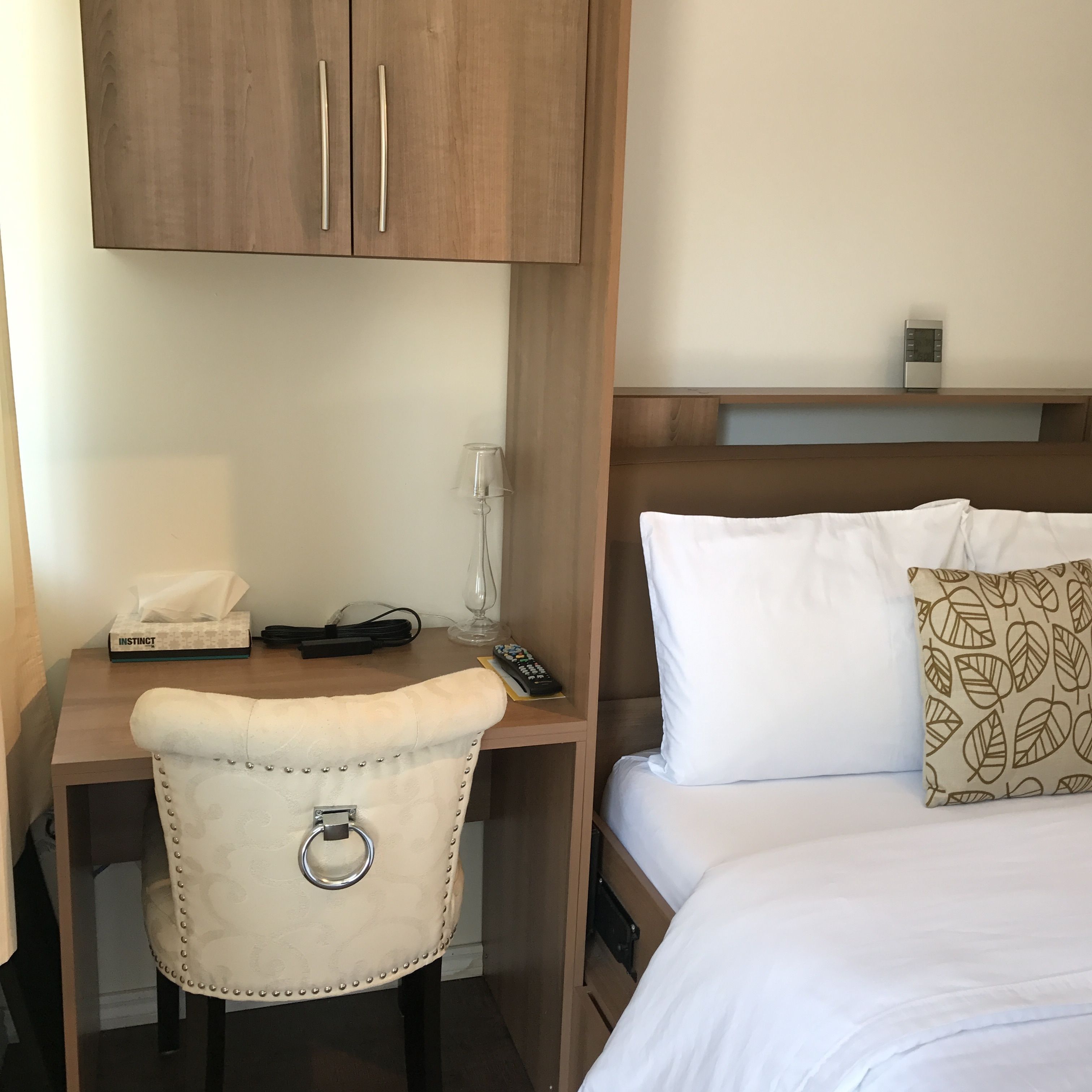 Quarto de hotel com uma secretária de madeira, uma cadeira branca com manta de pele, cama com roupa de cama branca e almofada estampada, e uma pequena prateleira com um controle remoto.
