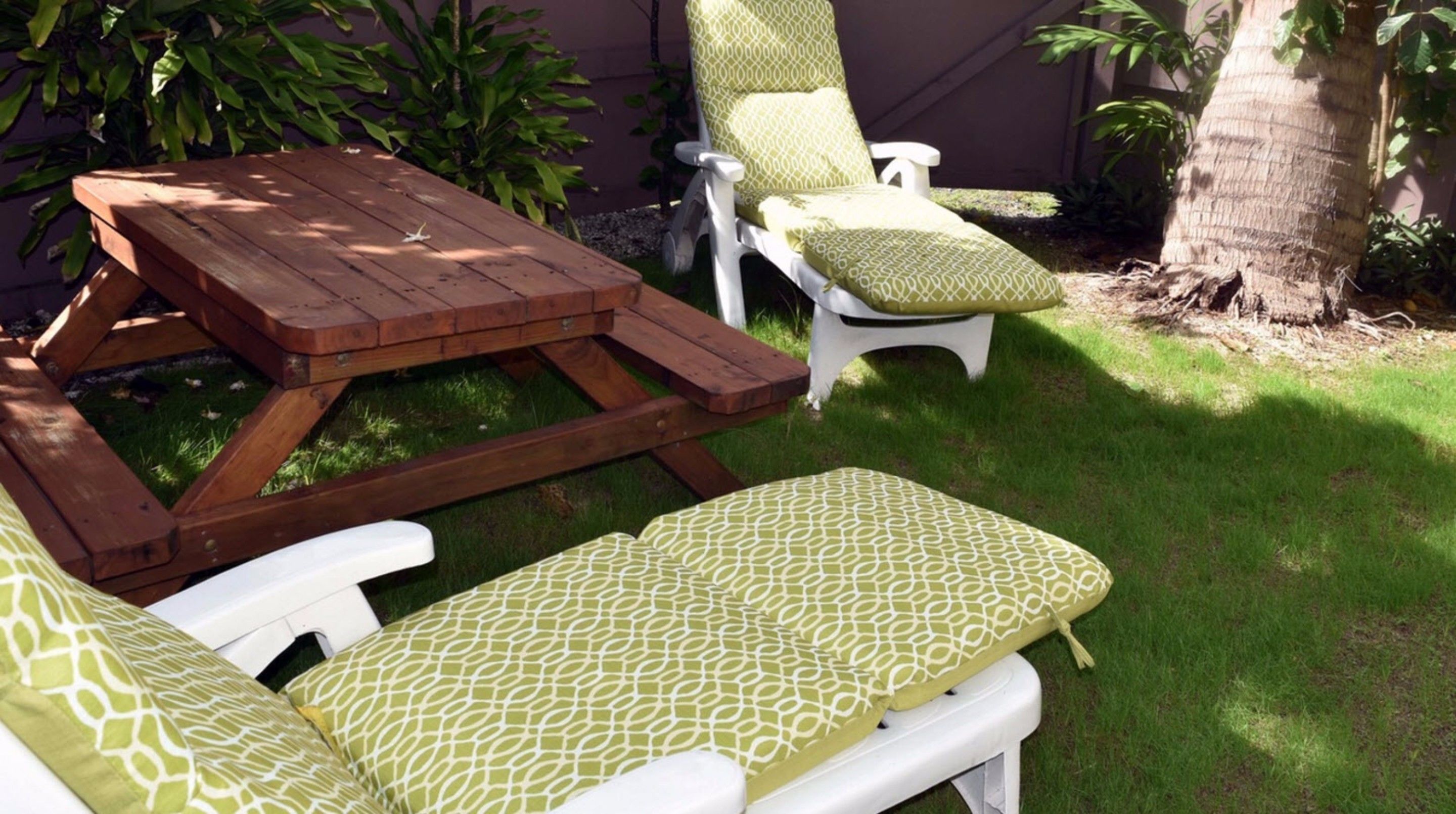 Uma mesa de piquenique de madeira e duas cadeiras de lounge brancas com almofadas estampadas em uma área gramada.