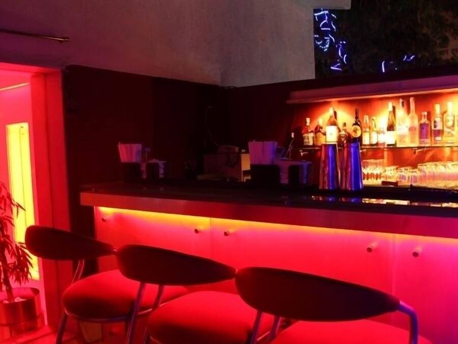 Uma área de bar com iluminação vermelha, um balcão com garrafas e três cadeiras vermelhas.