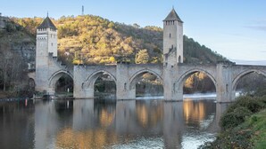 Uma ponte de pedra com duas torres sobre um rio calmo, rodeada por árvores e colinas.