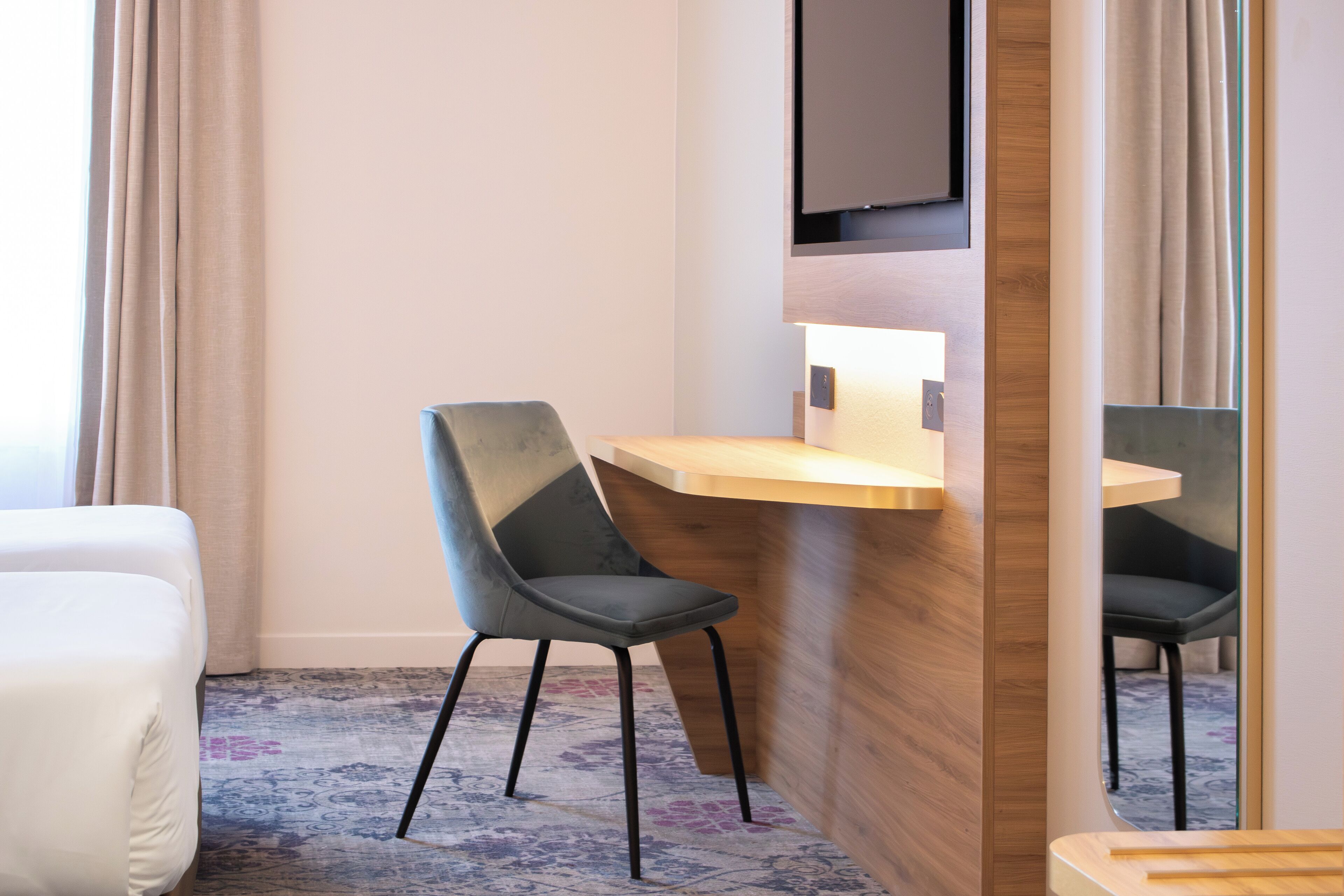 Quarto de hotel com cama, escrivaninha com cadeira, televisão de tela plana na parede e um espelho grande.