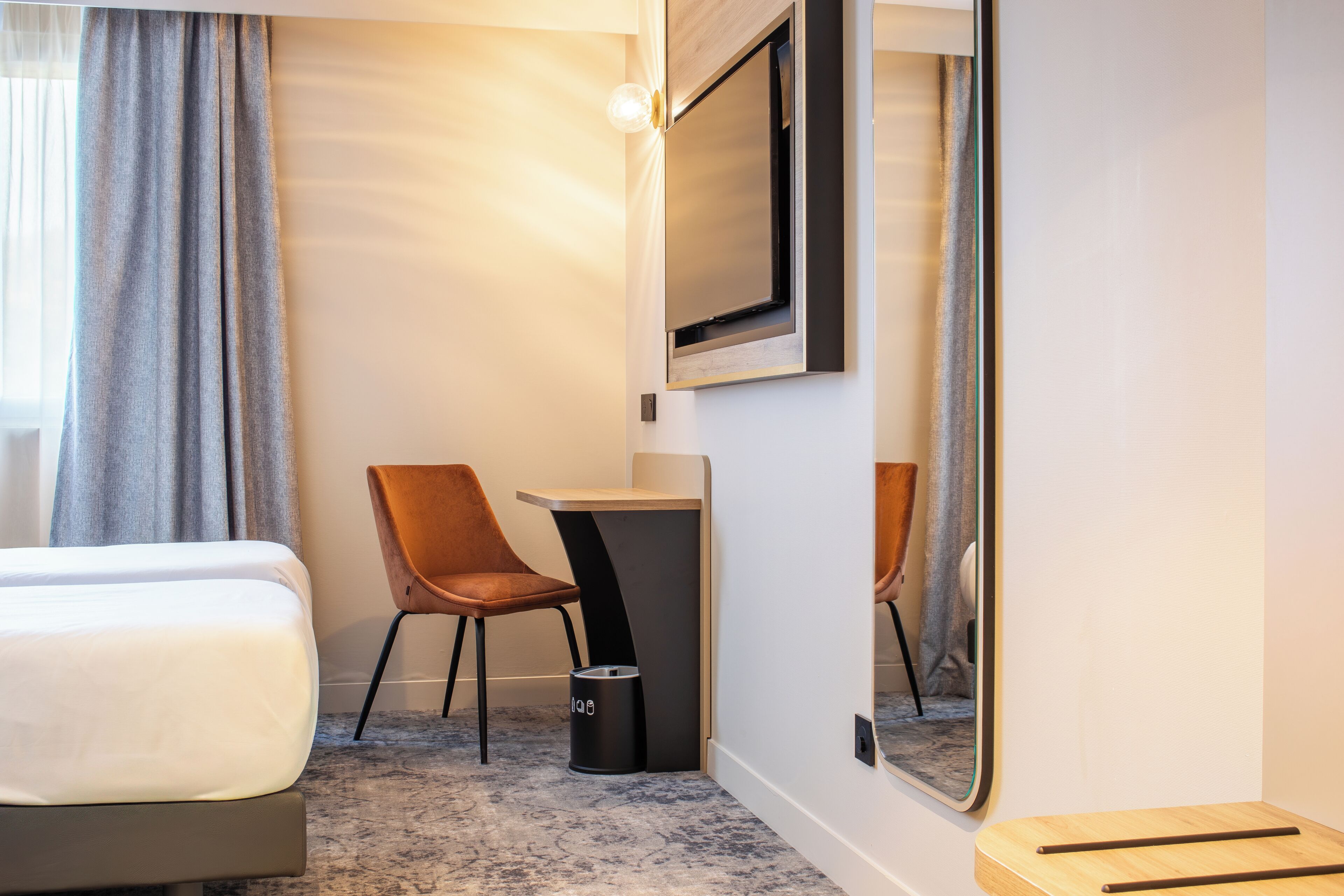 Quarto de hotel com cama, uma cadeira, mesa pequena, lixeira e espelho.