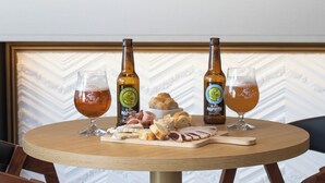 Uma mesa redonda com duas garrafas de cerveja e dois copos, um prato com doces, e uma cadeira de madeira. Ao fundo, uma parede com prateleiras exibindo garrafas.