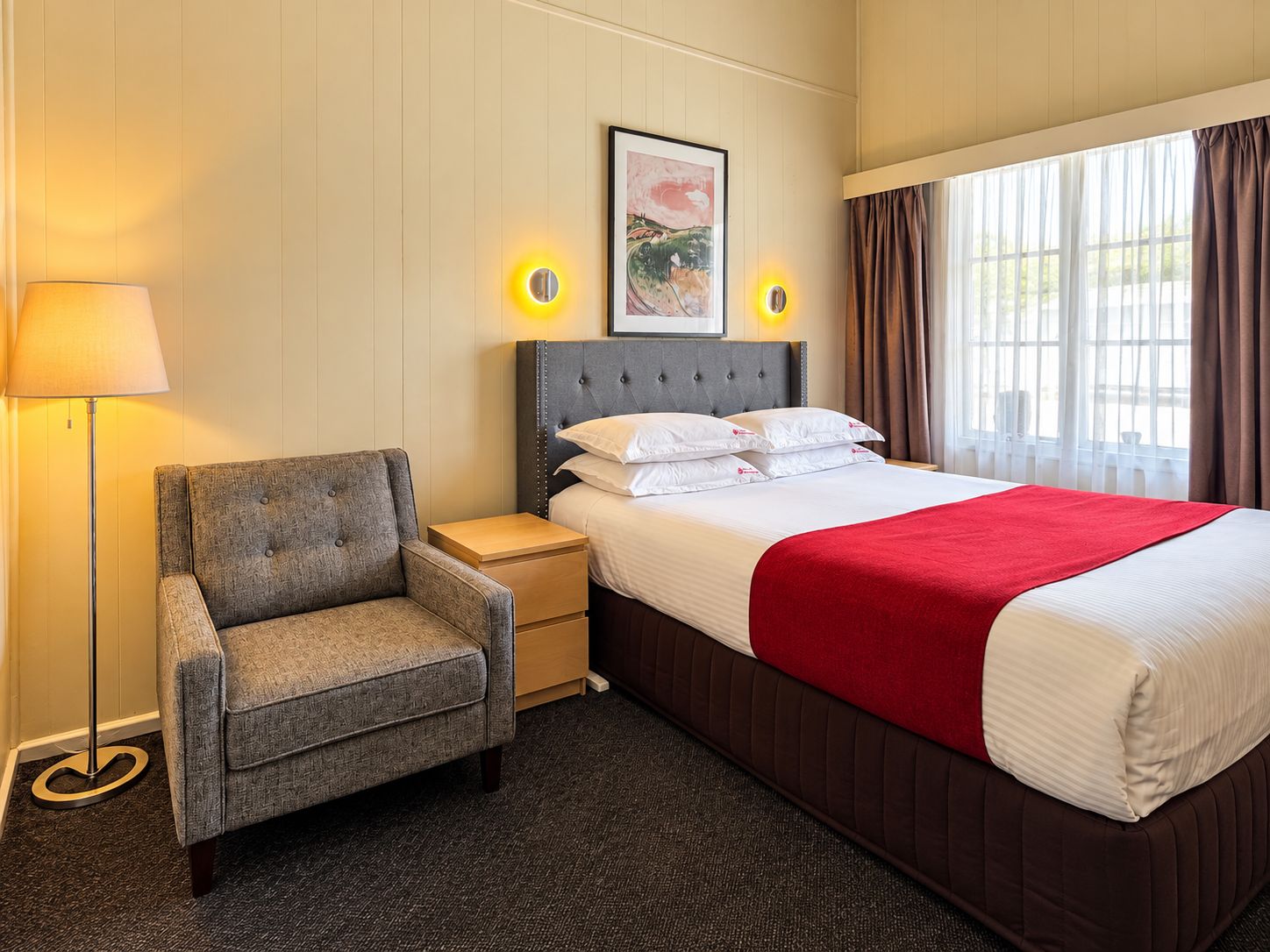 Quarto de hotel com cama, poltrona, mesa de cabeceira, abajur e um quadro na parede.